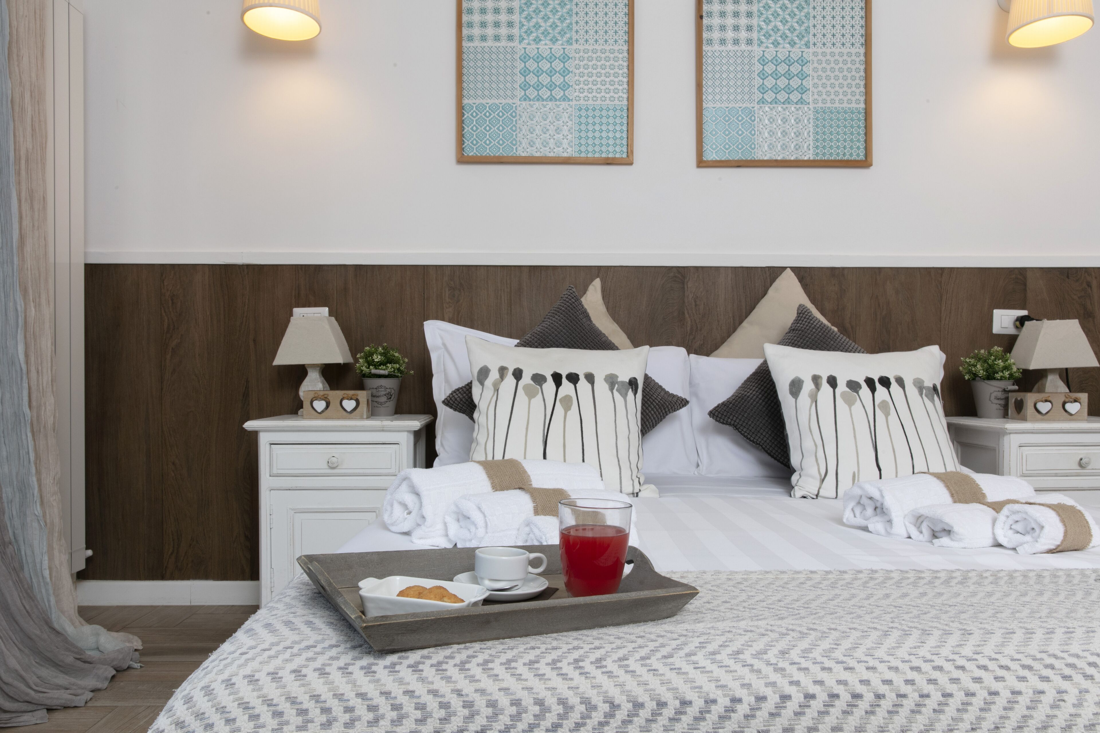 Cama bem arrumada com lençóis brancos, bandeja com uma xícara e um copo de bebida vermelha, e toalhas dobradas ao lado.
