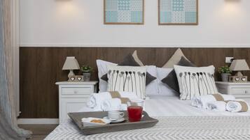 Cama bem arrumada com lençóis brancos, bandeja com uma xícara e um copo de bebida vermelha, e toalhas dobradas ao lado.