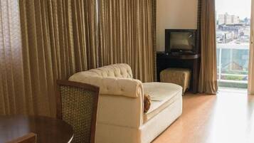 Quarto de hotel com sofá, televisão, mesa pequena e vista da cidade através de amplas janelas.