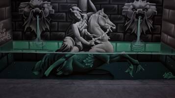 Escultura de uma mulher e um cavalo sobre uma plataforma verde, com um muro de tijolos ao fundo.