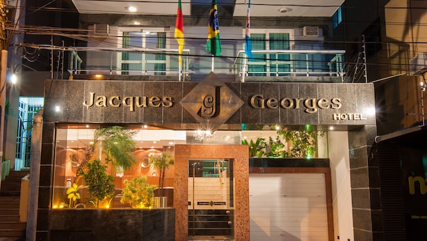 Entrada do Hotel Jacques Georges, com uma placa em destaque, fachada de vidro e área de acesso bem iluminada.