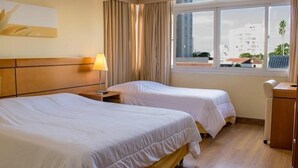 Quarto de hotel com duas camas, uma mesa de cabeceira com abajur, uma janela com cortinas e vista para prédios.