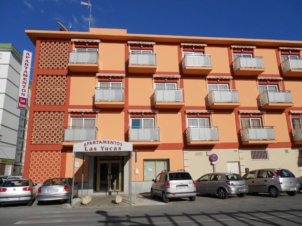 Um edifício de vários andares com varandas, um letreiro com a inscrição "Apartamentos Las Yucas" e vários carros estacionados na frente.