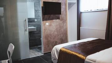 Quarto de hotel com cama, televisão na parede, banheiro com chuveiro e janela com cortinas.