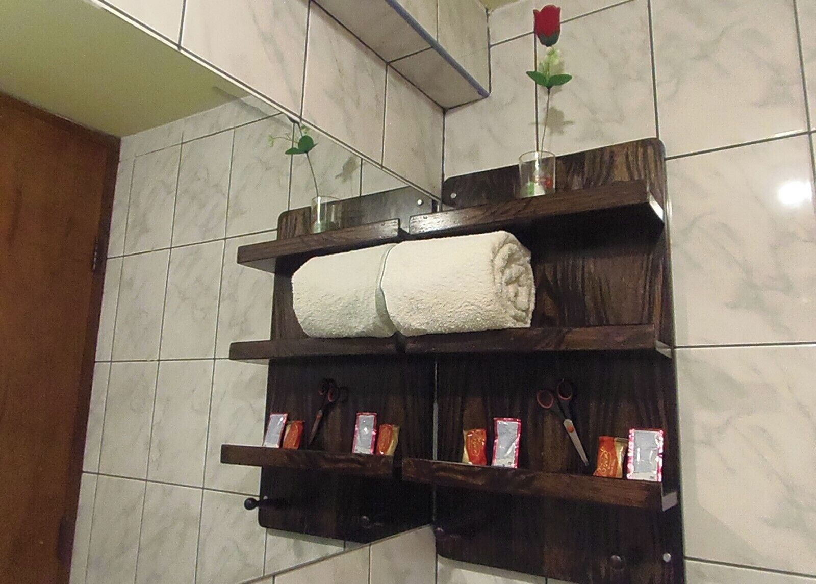 Casa de banho com parede revestida a mármore, prateleiras de madeira com toalhas e produtos de higiene, e uma rosa num vaso de vidro.