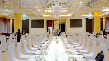 Uma sala de banquetes com longas mesas preparadas para um evento formal, cadeiras brancas e um candelabro.
