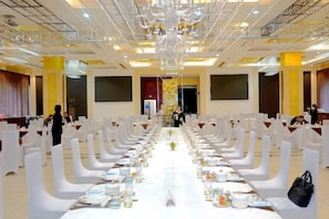 Uma sala de banquetes com longas mesas preparadas para um evento formal, cadeiras brancas e um candelabro.
