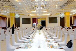 Uma sala de banquetes com longas mesas preparadas para um evento formal, cadeiras brancas e um candelabro.