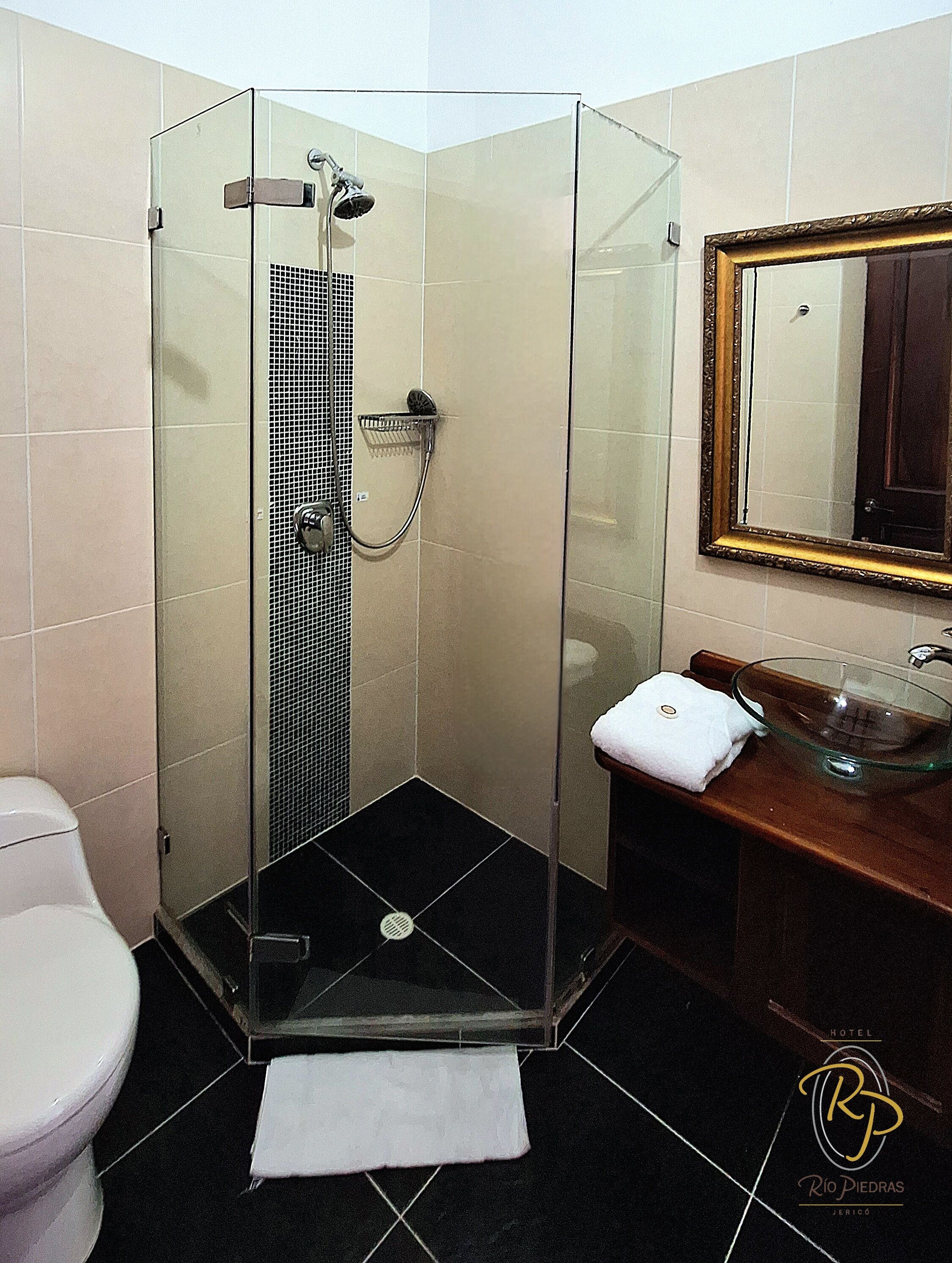 Um chuveiro com paredes de vidro, revestido com azulejos preto e branco, um vaso sanitário branco e um lavatório de madeira com cuba de vidro e uma toalha.