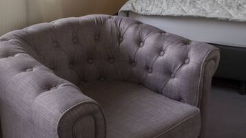Quarto com poltrona estofada, cama com cabeceira acolchoada, mesa de cabeceira com abajur e vigas de madeira no teto.