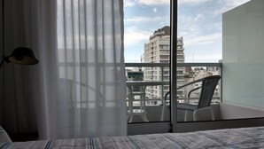 Quarto com cama, uma cadeira e vista para a cidade através de janelas amplas.