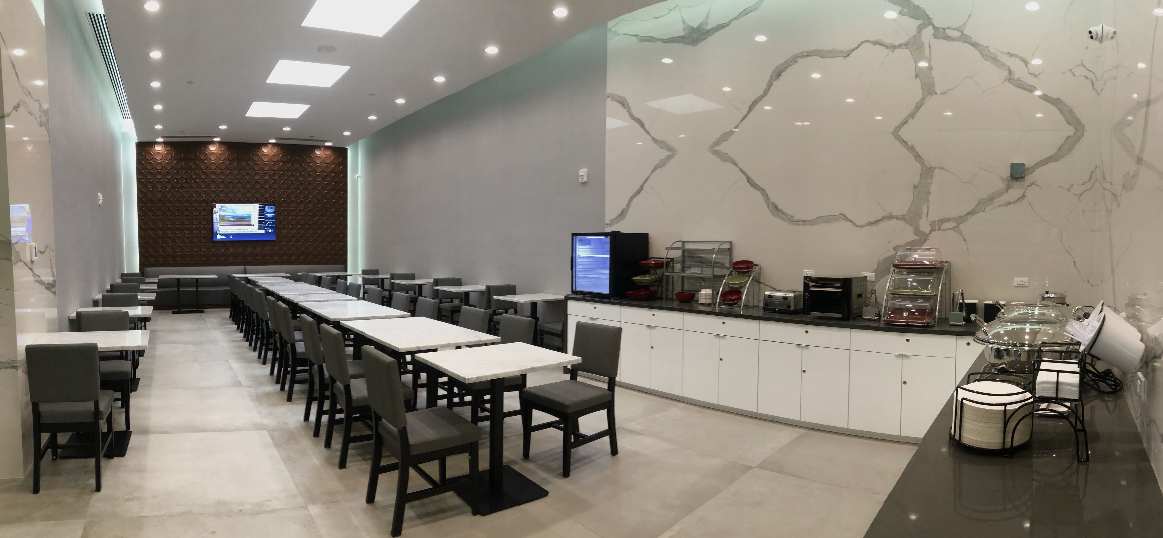Uma sala de jantar moderna com mesas e cadeiras compridas, uma bancada de cozinha com eletrodomésticos e uma tela de projeção na parede.