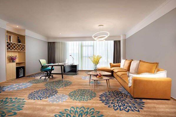 Quarto de hotel moderno com sofá, escrivaninha e cadeira. Possui uma janela grande com cortinas, tapete com padrão floral e luminária pendente.