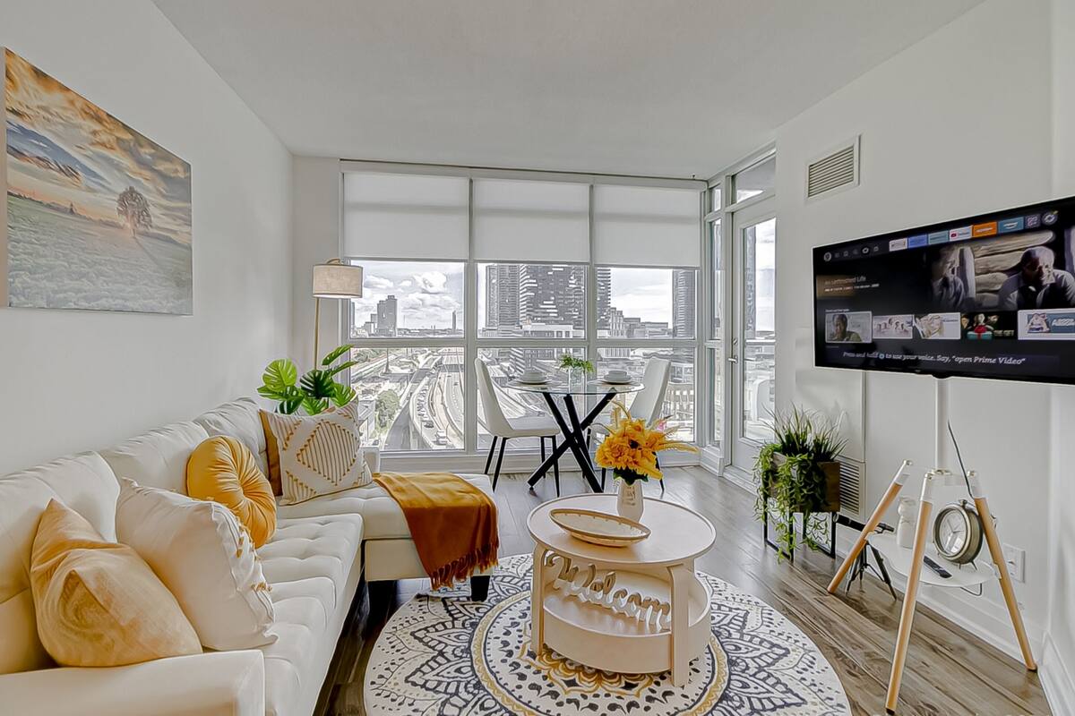 Uma sala de estar moderna com um sofá branco, uma mesa de centro com um vaso, uma TV na parede e uma janela grande com vista para a cidade.