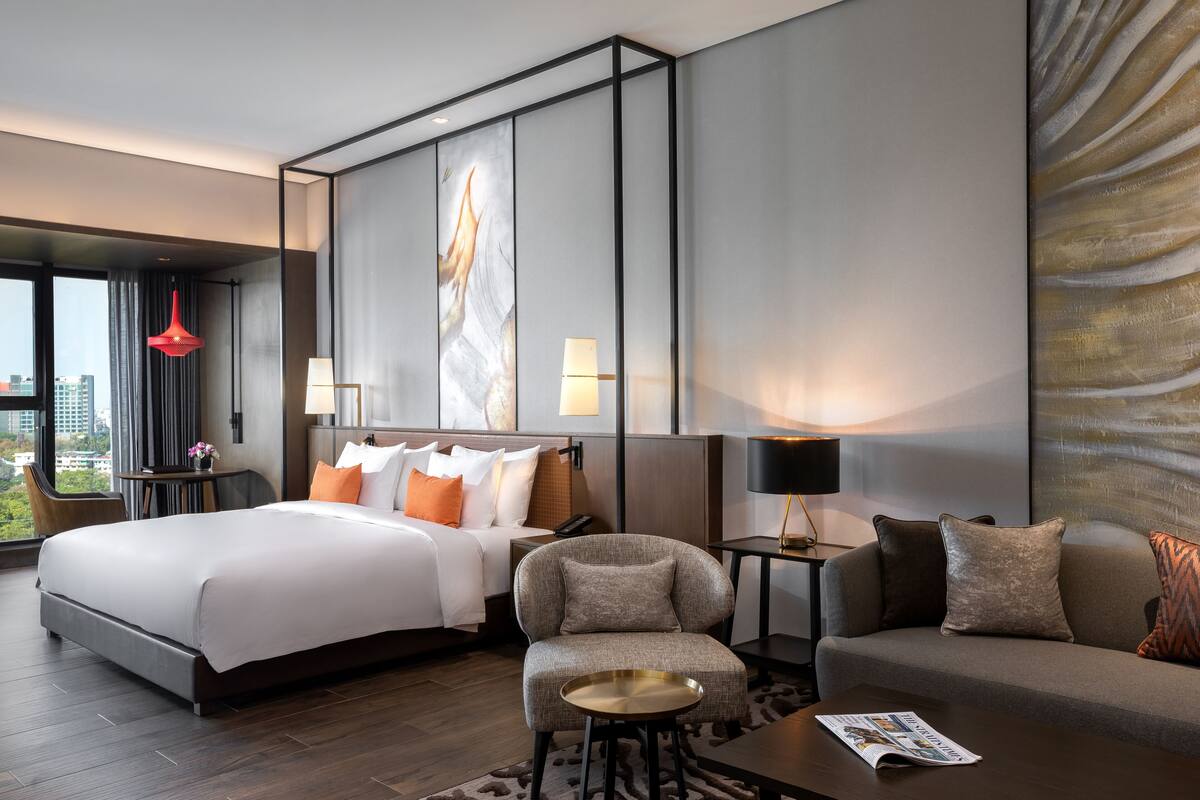 Quarto de hotel moderno com uma cama grande, um sofá, uma cadeira, uma mesinha e vista para a cidade.