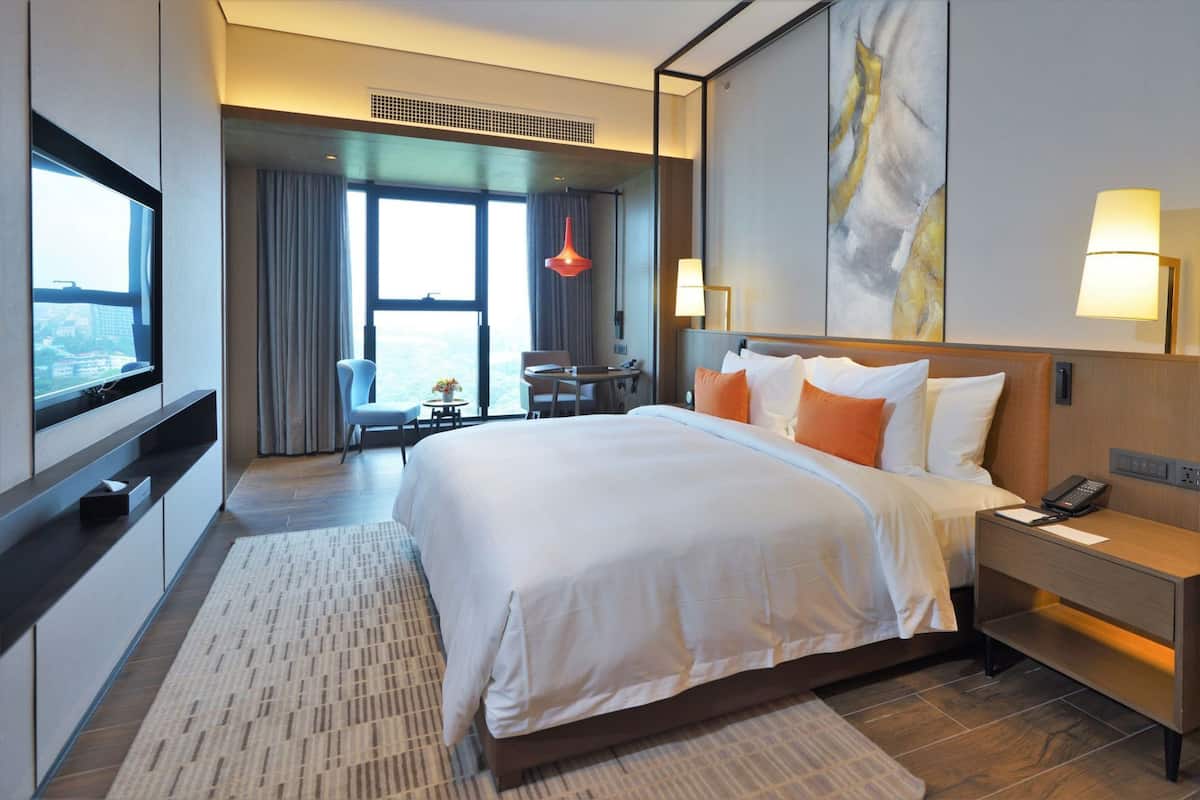 Quarto de hotel com uma cama grande, mesas de cabeceira, escrivaninha, cadeira, televisão e uma janela ampla com cortinas.