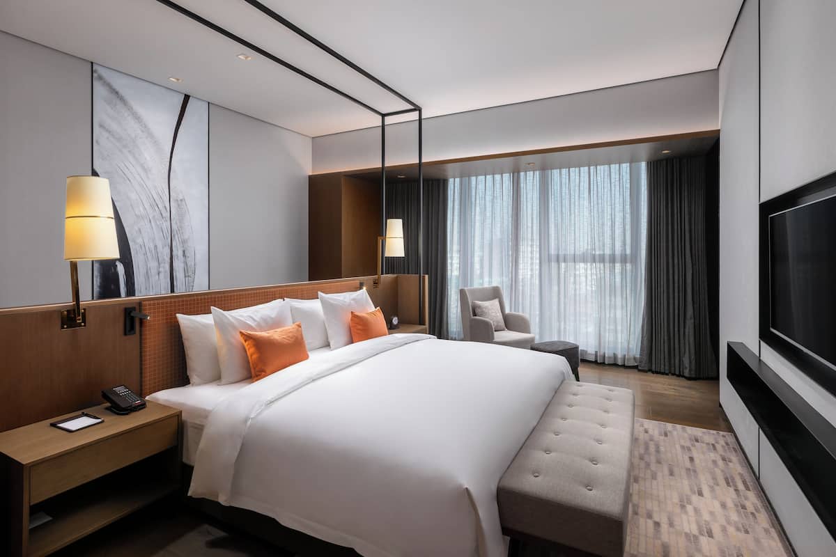 Quarto de hotel moderno com uma cama grande, uma escrivaninha com luminária, uma cadeira, uma televisão e uma janela ampla com cortinas.