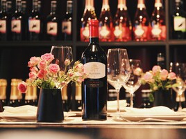Ambiente de bar com diversas garrafas de vinho, incluindo uma garrafa de vinho Badagoni, e vasos com rosas cor-de-rosa.