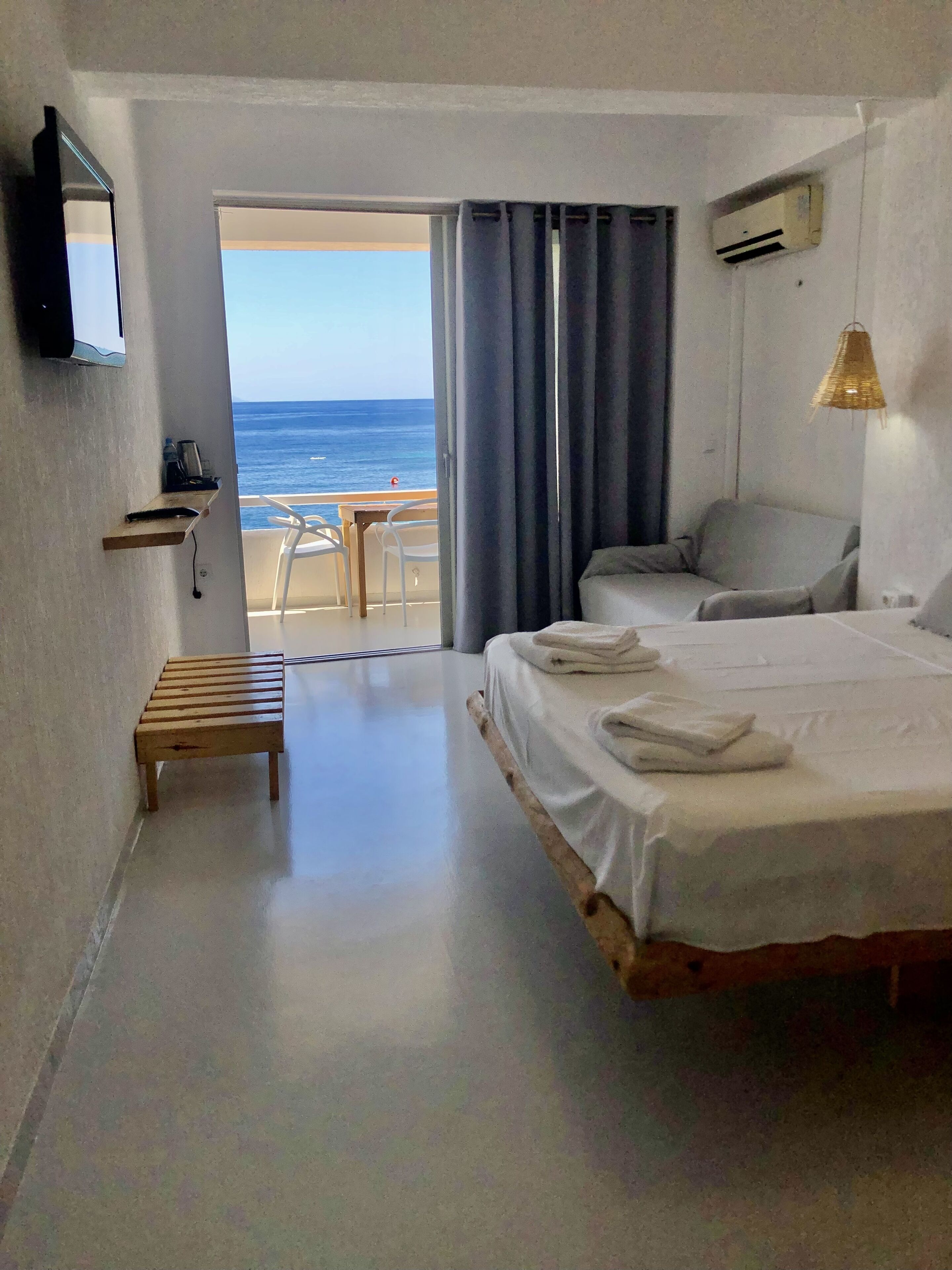 Quarto de hotel com cama, uma mesinha, uma cadeira, TV fixada na parede e vista para o mar.