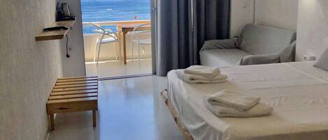 Quarto de hotel com cama, uma mesinha, uma cadeira, TV fixada na parede e vista para o mar.