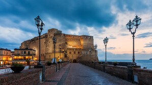 Uma fortaleza de pedra histórica com um caminho de paralelepípedos que leva à sua entrada, ladeada por postes de luz e vista para a orla marítima.