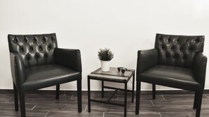 Duas poltronas de couro preto com design acolchoado, dispostas de cada lado de uma pequena mesa de madeira com uma planta em vaso e óculos de sol.