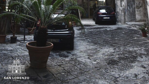 Um pátio urbano com um arco de pedra, plantas em vasos e um carro estacionado.
