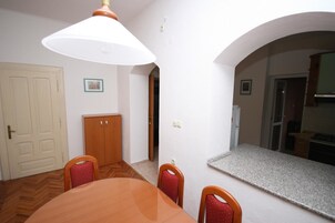 Uma sala de jantar com uma mesa e cadeiras de madeira, uma luminária branca no teto e uma cozinha visível através de um arco.