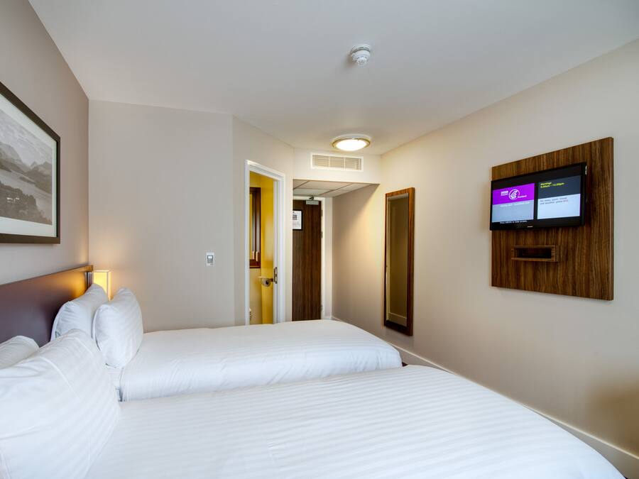 Quarto de hotel com duas camas, uma TV fixada na parede, um quadro enquadrado e uma luminária no teto.