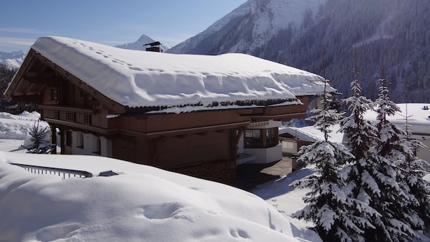 Um chalé com telhado coberto de neve, rodeado por árvores e montanhas cobertas de neve.