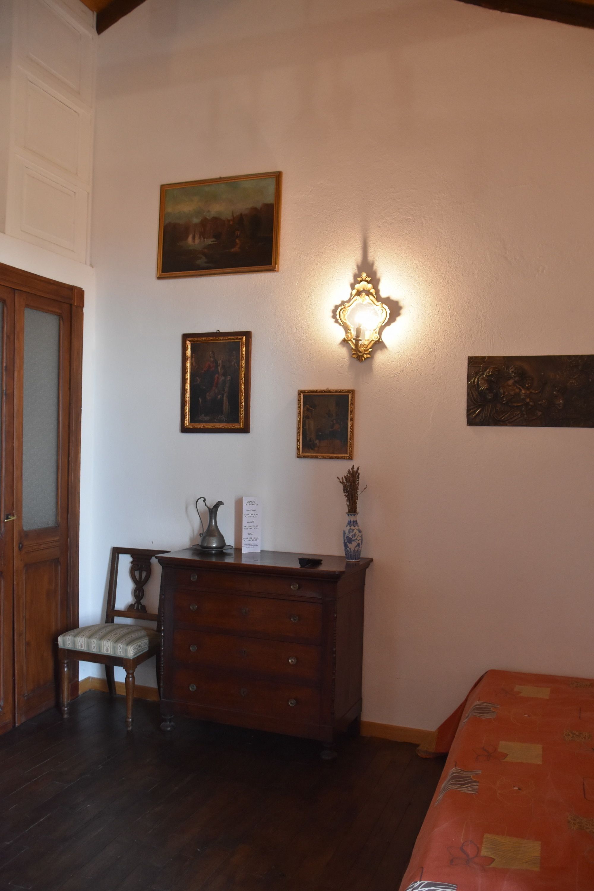 Quarto com cômoda de madeira, uma cadeira e uma cama. Há três quadros na parede e uma luminária fixada.