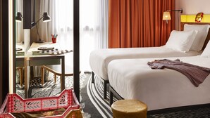 Quarto de hotel com uma cama, uma escrivaninha, uma cadeira, um abajur e um banquinho pequeno.