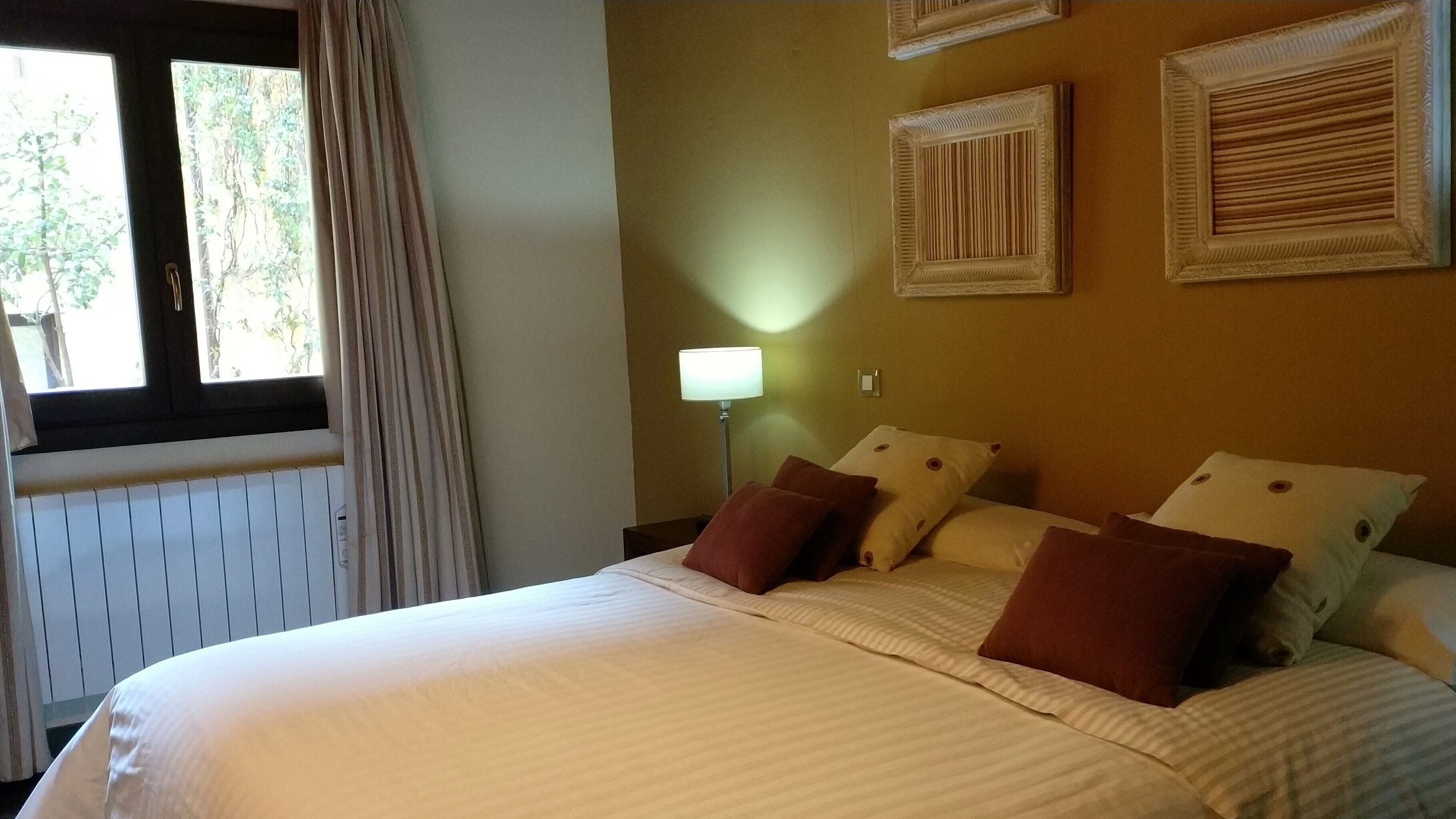 Quarto de hotel com uma cama grande, janela com cortinas, luminária de pé e obras de arte emolduradas na parede.