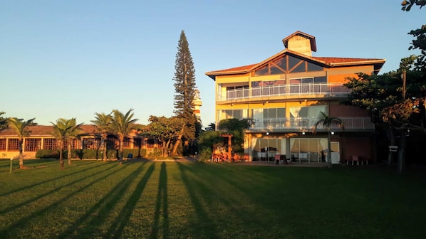 Um edifício de dois andares com varanda, rodeado por um gramado bem cuidado e palmeiras.