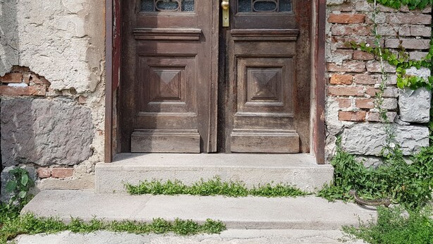 Uma porta dupla de madeira com detalhada ferragem metálica e um pequeno degrau que a precede.