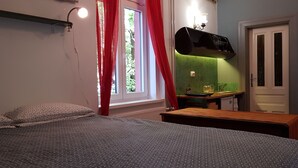 Quarto com cama, uma mesa pequena, um banco, janela com cortinas e porta.