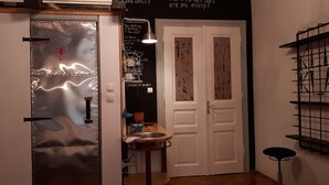 Um corredor estreito com uma porta de metal à esquerda, uma porta branca à direita e uma pequena mesa com uma tigela e um pano.