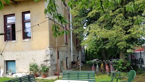 Uma área de estar ao ar livre com mesas e cadeiras de madeira, cercada por vegetação e um edifício com janelas.