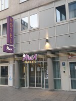 Entrada de hotel com um letreiro chamativo 'Awa Hotel', portas de vidro e uma placa de rua indicando 'Schillerstr. 10'.