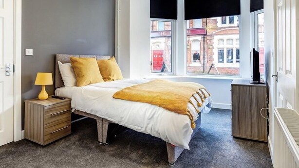 Quarto com cama, mesa de cabeceira, abajur e vista da rua pela janela.