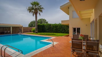 Piscina com deck revestido, uma palmeira e área de varanda com mobiliário externo.