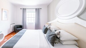 Quarto de hotel moderno com uma cama grande, uma mesinha de cabeceira, uma cadeira e uma janela com cortinas.