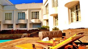 Uma área à beira da piscina com espreguiçadeiras e uma pequena mesa, cercada por um edifício com janelas amplas e uma parede de tijolos.