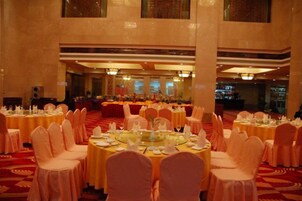 Uma sala de banquetes com mesas redondas dispostas para refeições, pé-direito alto e vista para uma área de bar ao fundo.