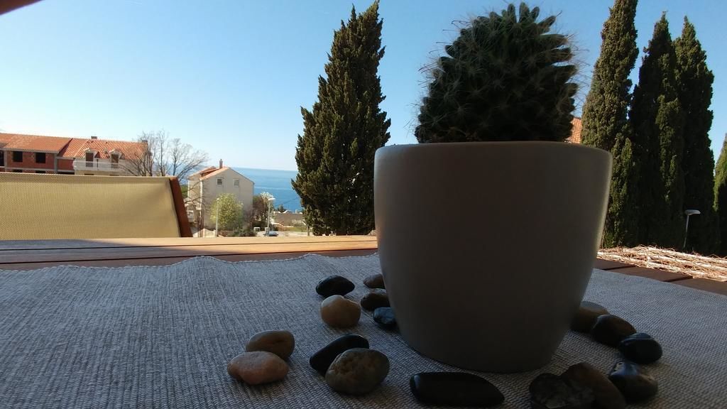 Vista de uma varanda com uma tigela branca grande sobre um tapete, cercada por pedras, com vista para uma área costeira com edifícios e árvores altas.
