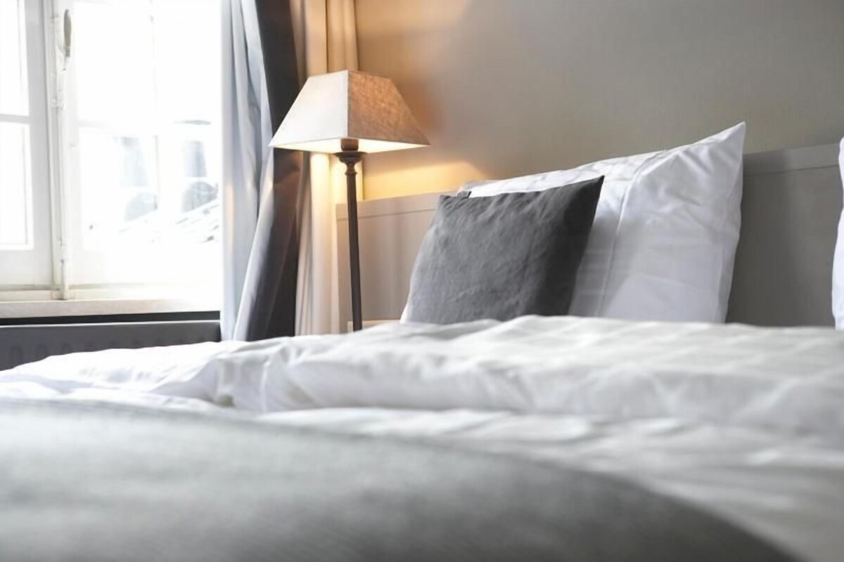 Quarto de hotel com cama, luminária e janela com cortinas.