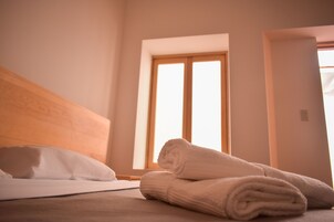Cama bem arrumada com lençóis brancos e toalha enrolada, cabeceira de madeira e janela com persianas verticais em um quarto de paredes claras.