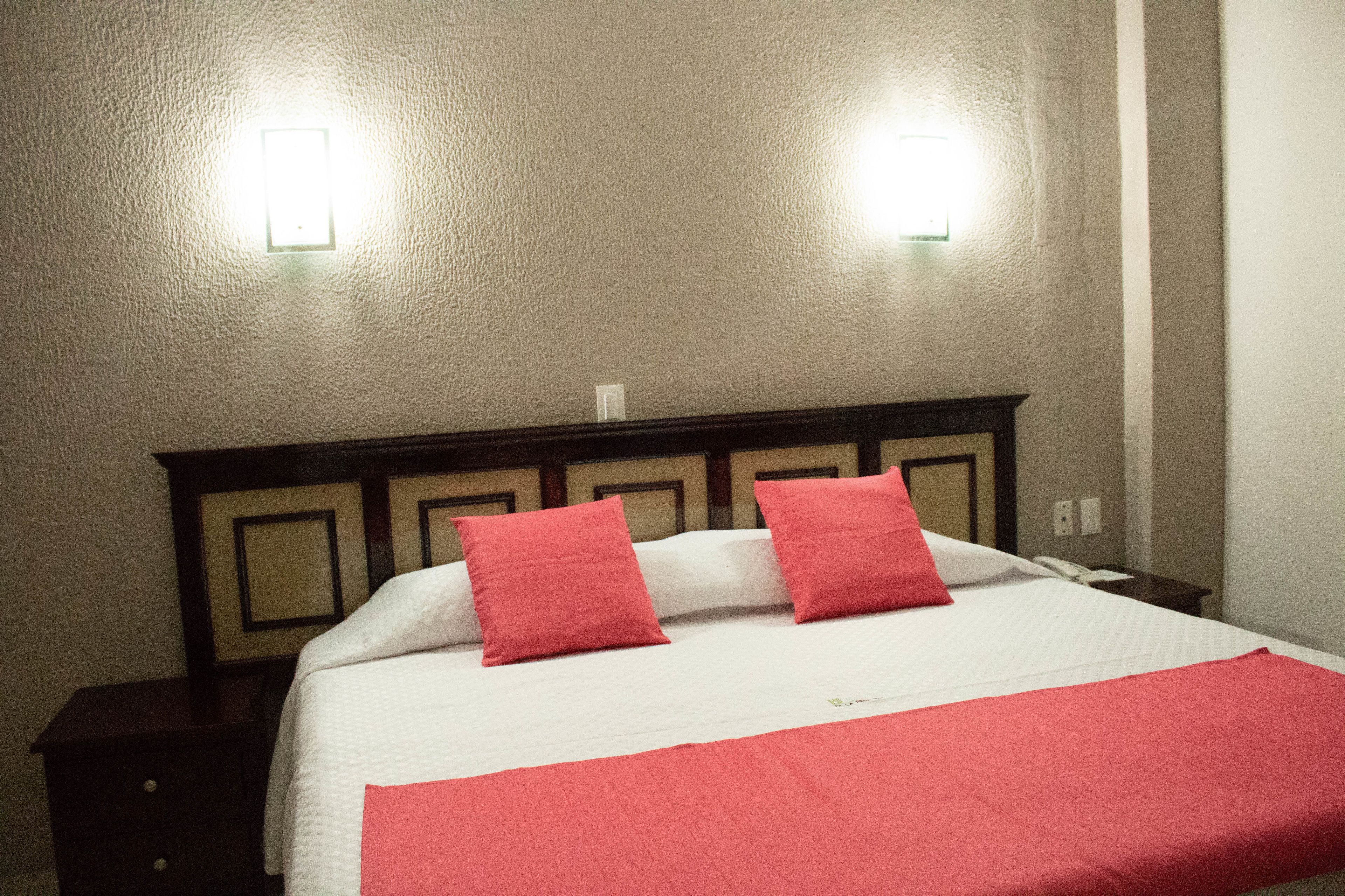 Quarto de hotel com uma cama, dois travesseiros vermelhos, um edredom branco e duas luminárias na parede.