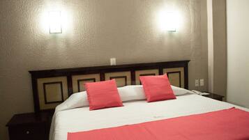 Quarto de hotel com uma cama, dois travesseiros vermelhos, um edredom branco e duas luminárias na parede.