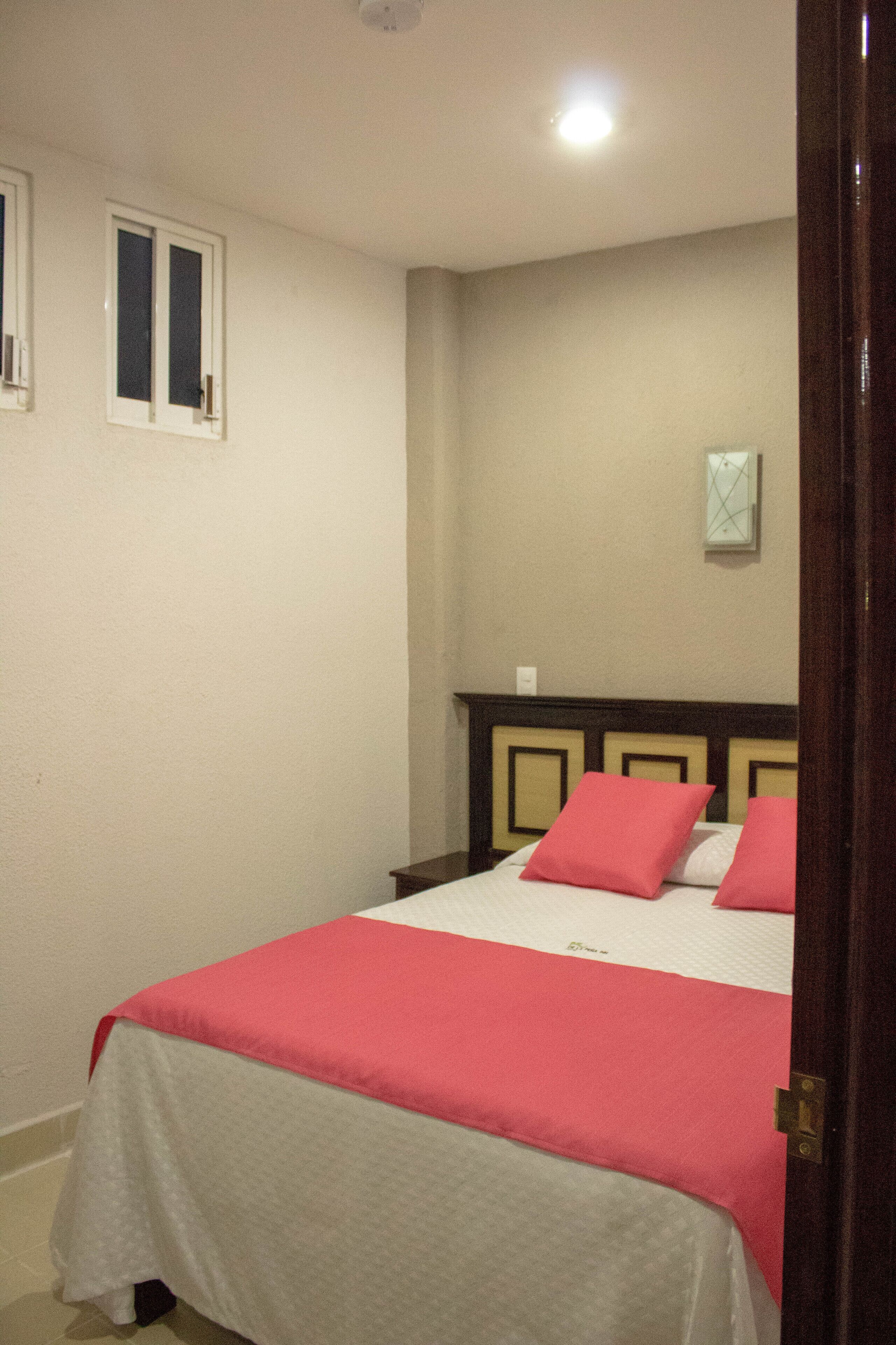 Quarto com cama, mesa de cabeceira, janela e luminária fixada na parede.